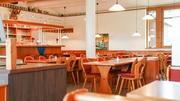 Interior de restaurante com mesas e cadeiras de madeira, área de bar com prateleiras cheias de copos, e iluminação pendente.
