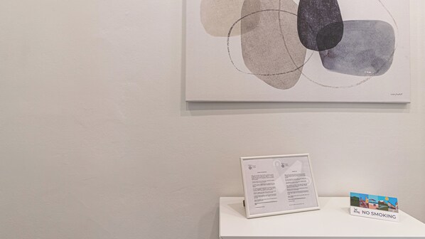 Uma obra de arte abstrata emoldurada numa parede, uma placa com texto, e um sinal de 'Não Fumar' sobre uma mesa.