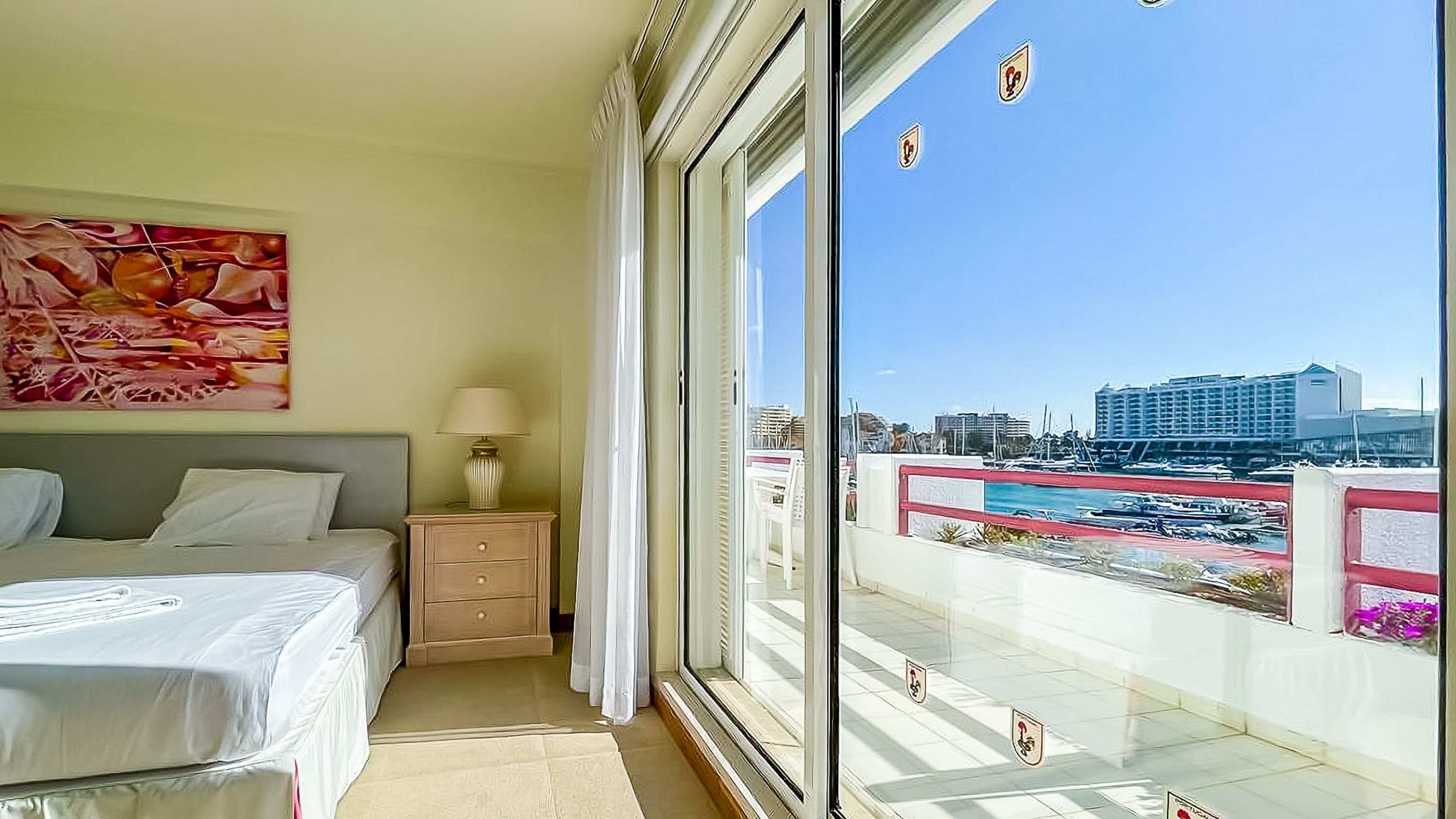 Quarto de hotel com cama, mesa de cabeceira, luminária e uma grande janela com vista para um porto e edifícios.