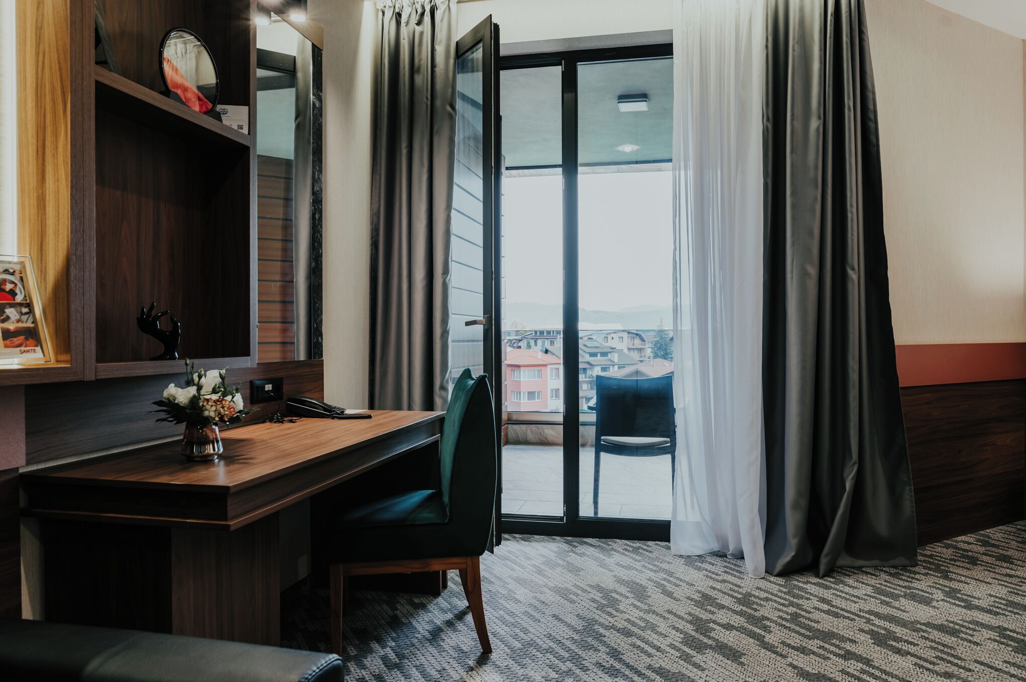 Quarto de hotel com uma secretária de madeira, uma cadeira, uma mesinha com um vaso de flores, um espelho e vista da cidade através de uma grande porta de correr de vidro.