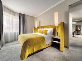 Quarto de hotel moderno com uma cama grande, mesa de cabeceira, abajur e vista da cidade pela janela.