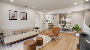 Uma sala de estar moderna com sofá, poltrona e mesa de centro. Há uma área de jantar com mesa e cadeiras. O cômodo tem paredes brancas e piso de madeira.