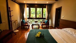 Quarto de hotel com cama, mesa de jantar com cadeiras, um ventilador e vista para o verde lá fora.