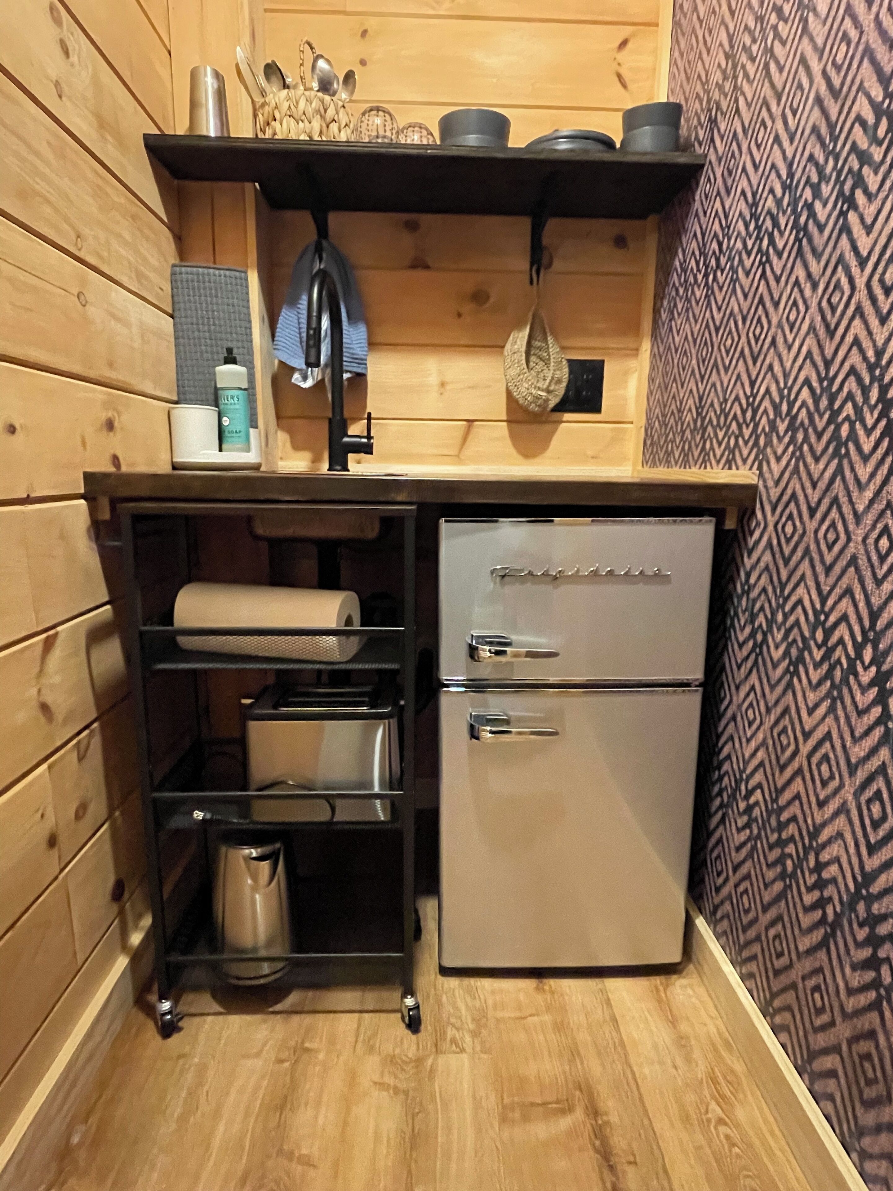Uma cozinha compacta com geladeira de aço inoxidável, pia e prateleira com itens básicos de cozinha.