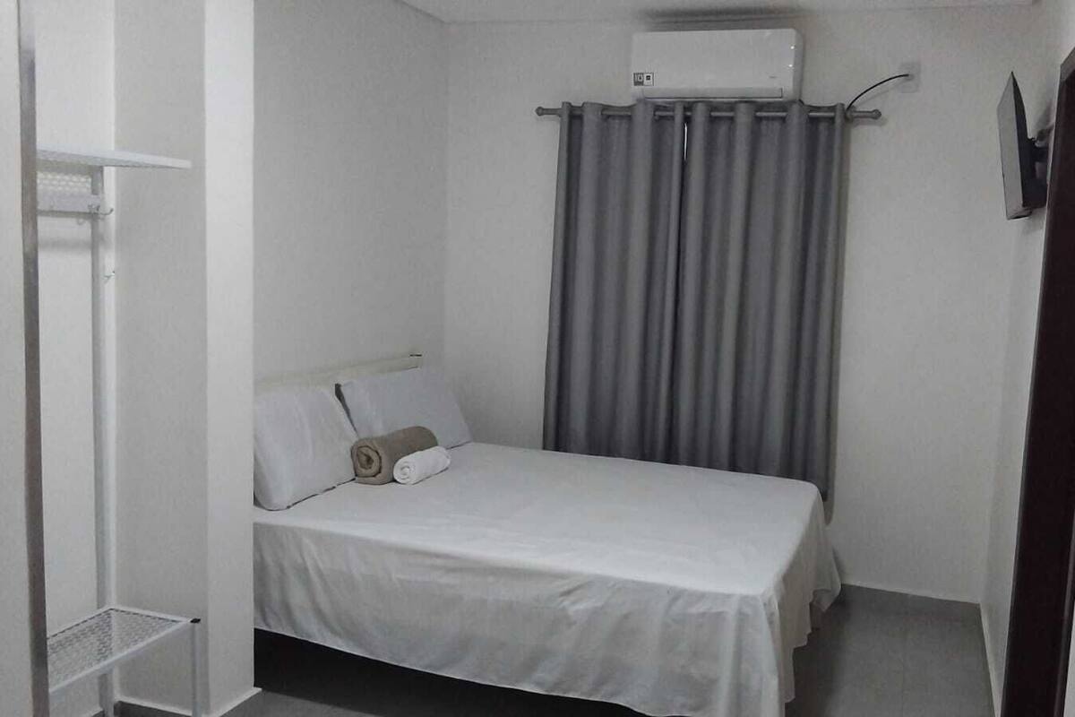 Quarto pequeno com uma cama de solteiro, colcha branca, cortina cinza e radiador branco sobre a cama.