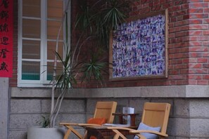 Edifício de tijolos com uma janela exibindo uma colagem de fotografias. Dois cadeiras de madeira e uma mesa estão do lado de fora.