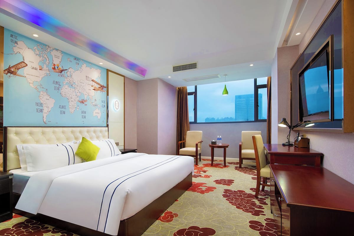 Quarto de hotel com um grande mapa-múndi na parede, cama com roupa de cama branca, escrivaninha com abajur e vista para a cidade pela janela.