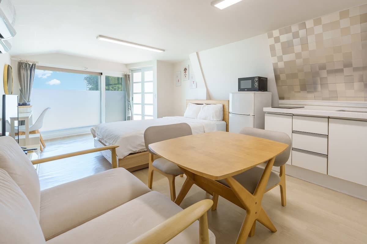 Quarto de hotel moderno com cama, mesa de jantar com cadeiras, cozinha compacta e uma janela grande.