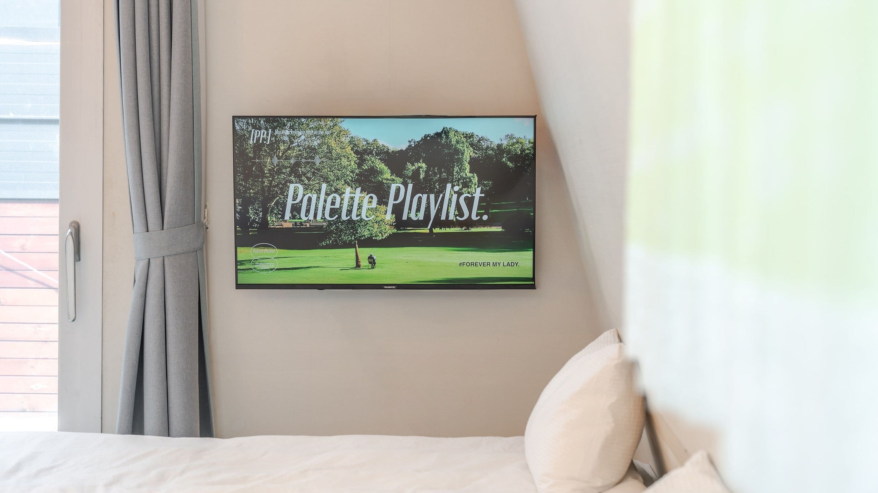 Quarto com uma cama, uma TV na parede exibindo uma 'Palette Playlist' com uma cena de parque e uma janela com cortinas.
