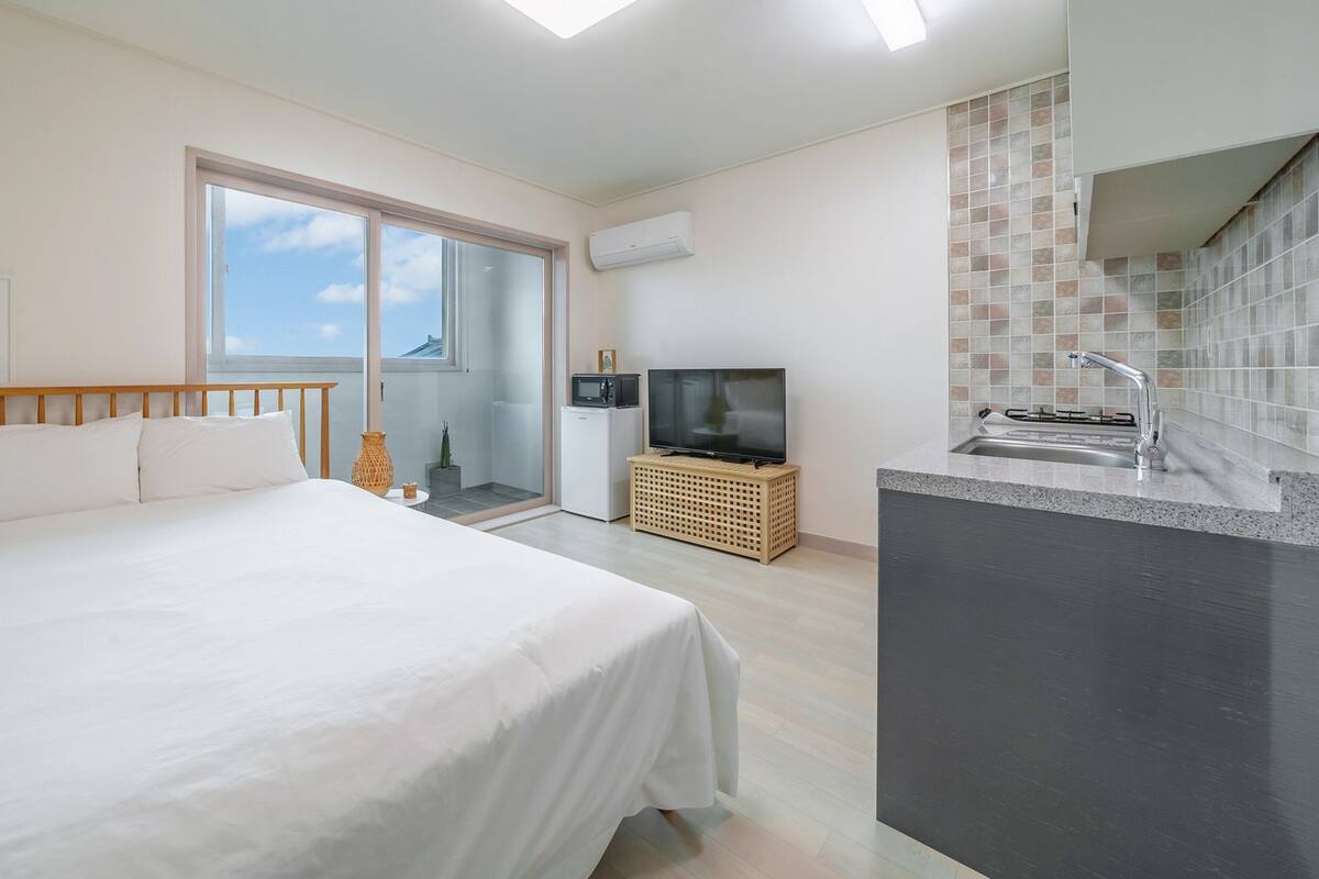 Quarto com cama, televisão, pequeno espaço de cozinha e porta de vidro deslizante para uma varanda.