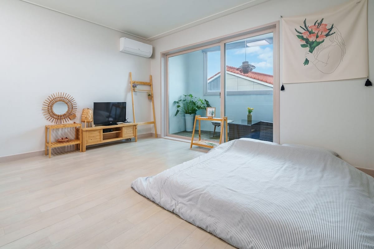 Quarto moderno e bem iluminado com uma cama grande, uma televisão sobre um suporte de madeira, uma mesinha e uma porta de correr de vidro que dá para uma varanda com um vaso de planta.