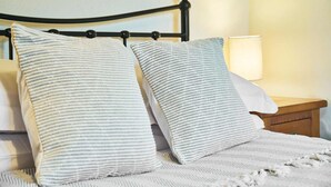 Uma cama com roupa de cama listrada e duas almofadas, uma mesa de cabeceira de madeira com um abajur e uma parede lisa.