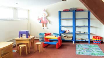 Uma sala de jogos infantil com uma mesa de madeira, banquetas, um tapete de atividades, uma estante azul e caixas organizadoras.