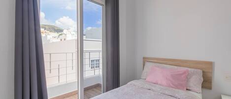Quarto com cama, janela com cortinas, vista de prédios e uma mesinha de cabeceira.