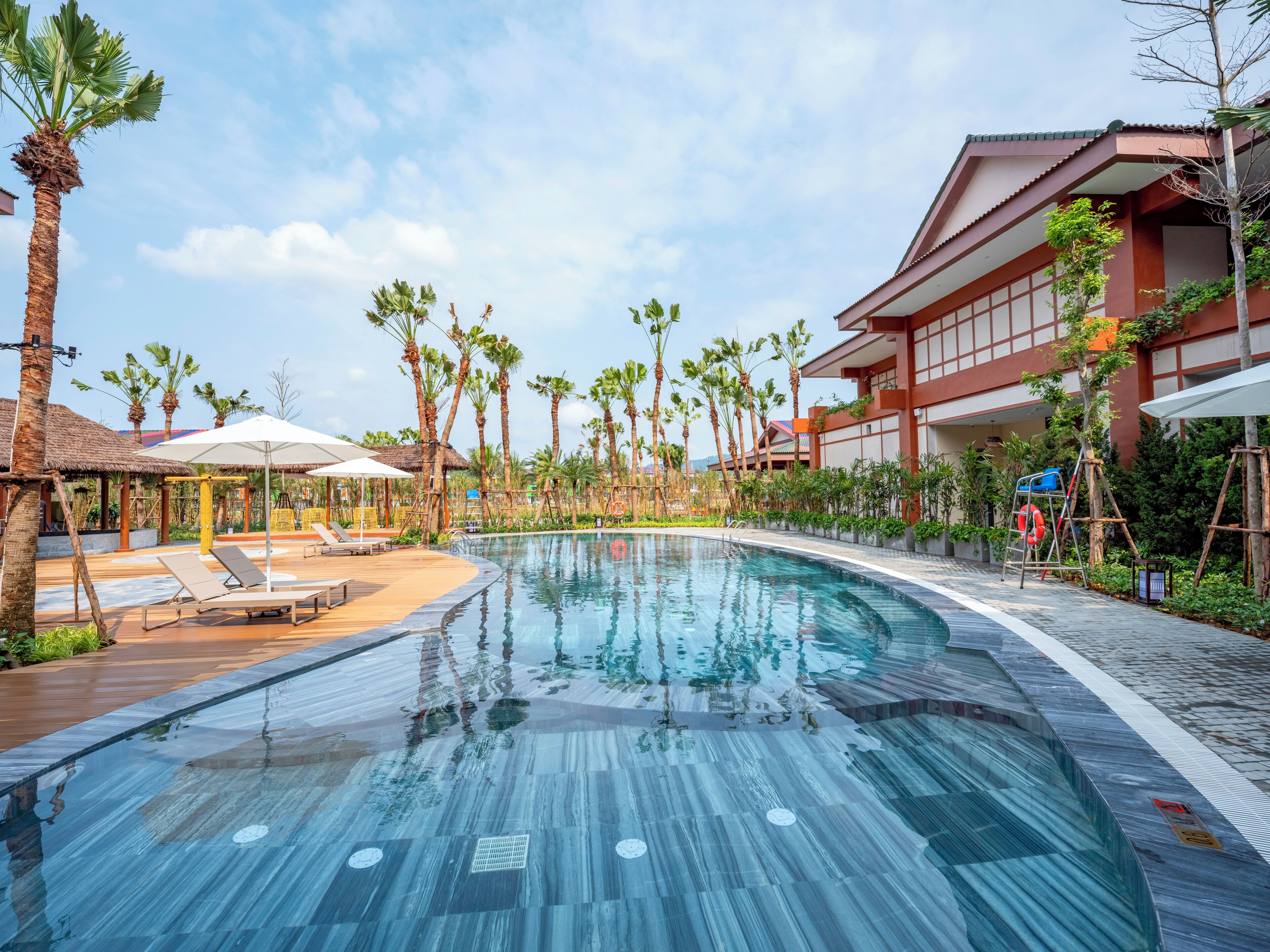 Piscina com espreguiçadeiras e guarda-sóis, cercada por palmeiras e um edifício com fachada vermelha e branca.