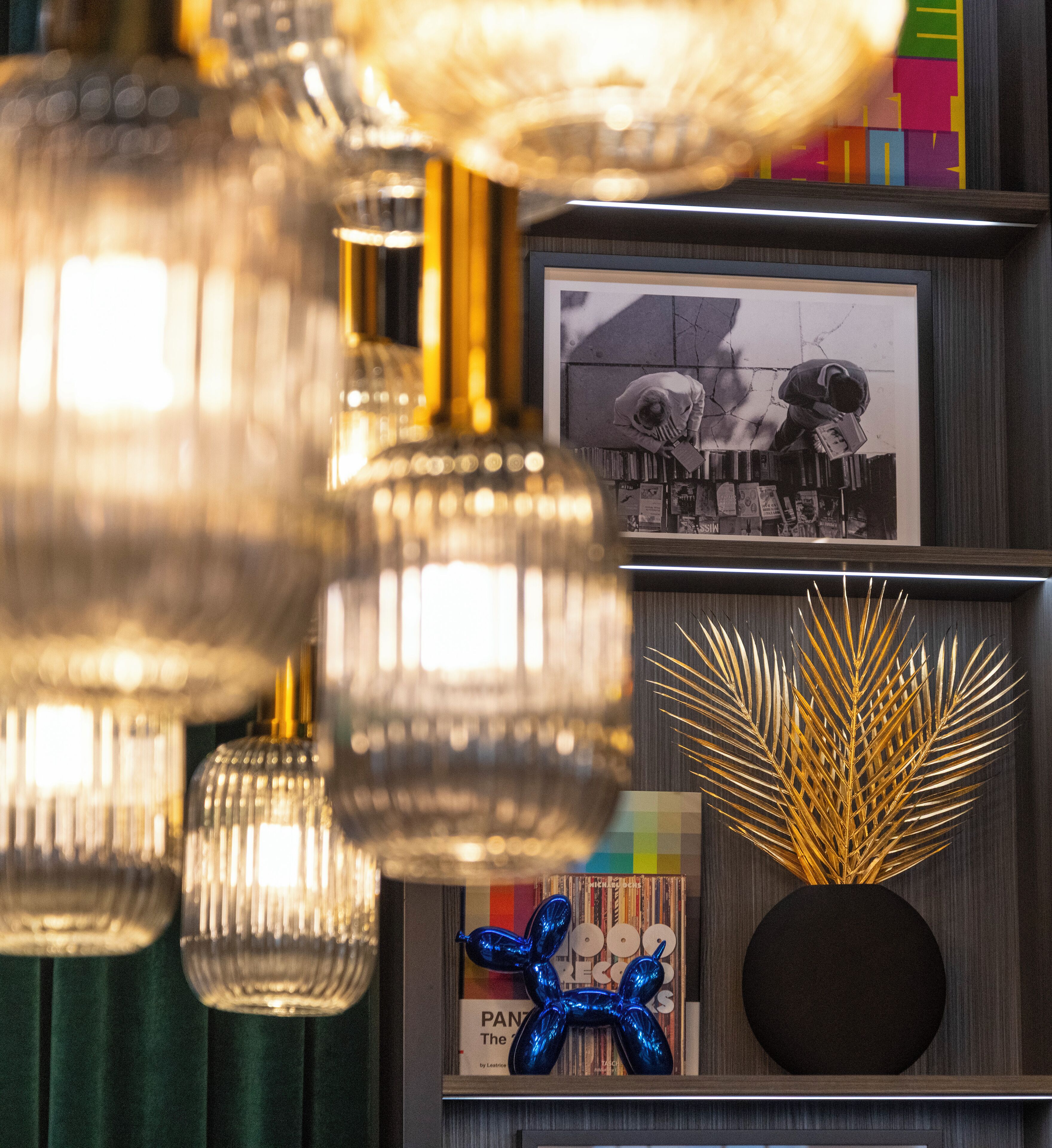 Uma parede com uma fotografia em preto e branco emoldurada, um frond de palmeira dourado decorativo e uma escultura de balão azul.