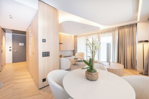 Quarto de hotel moderno com mesa de jantar redonda, assentos confortáveis, uma planta em vaso, sofá, pequena cozinha e uma grande janela com cortinas.