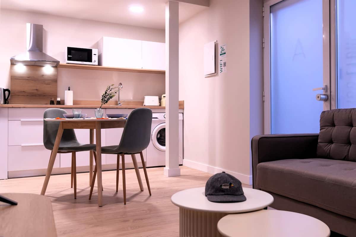 Uma sala de estar moderna com um sofá e um espaço de jantar com mesa e cadeiras. Há uma máquina de lavar e um micro-ondas na cozinha.