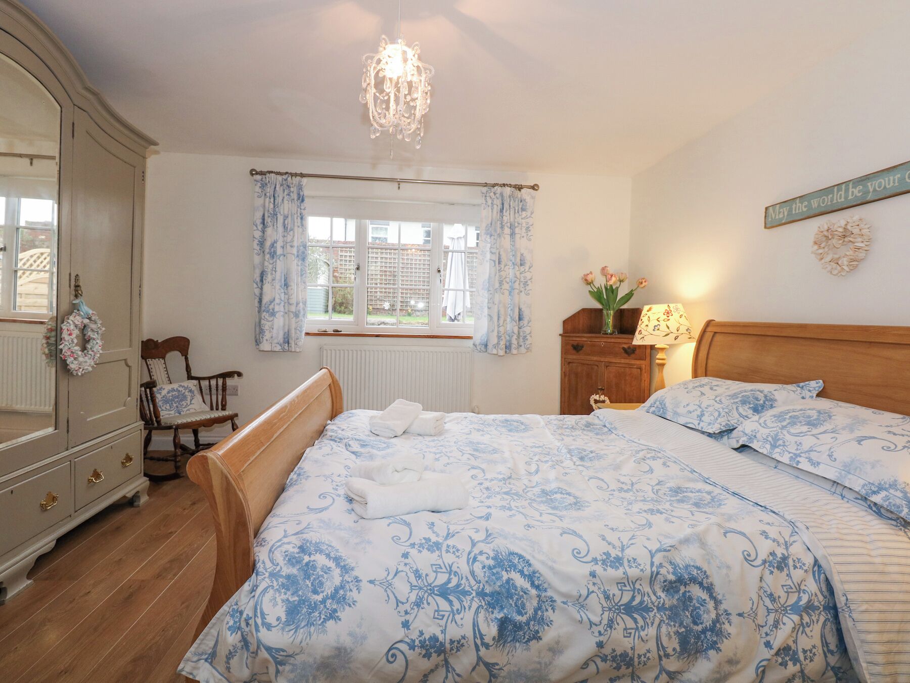 Quarto com cama de madeira, penteadeira, cadeira, janela com cortinas, lustre e decoração na parede.
