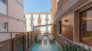 Um corredor estreito com piso de azulejos, uma área de estar coberta com cadeiras e vista para edifícios próximos.