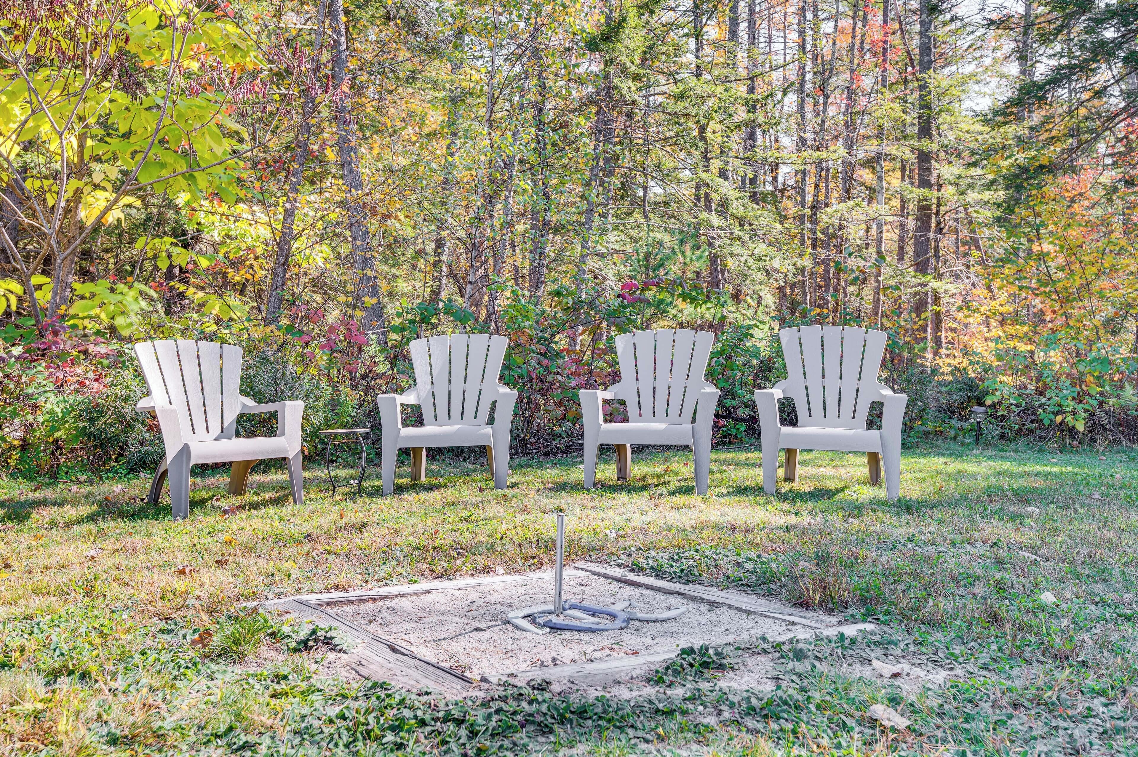 Três cadeiras Adirondack brancas num jardim com uma lareira no centro.