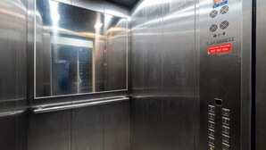 Interior de elevador com paredes metálicas, um painel de vidro refletivo e um painel de comando com botões e visor.