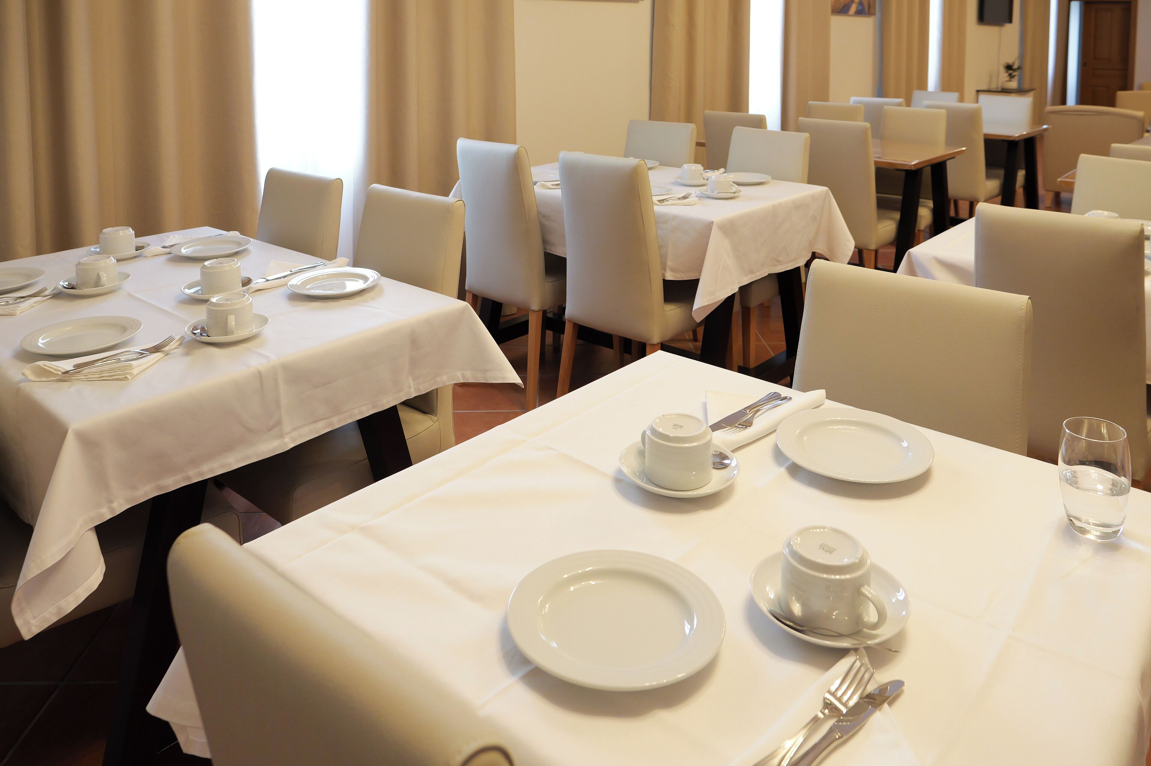 Uma sala de jantar com mesas preparadas para o serviço, com toalhas brancas e cadeiras.