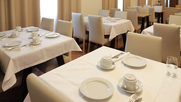 Uma sala de jantar com mesas preparadas para o serviço, com toalhas brancas e cadeiras.