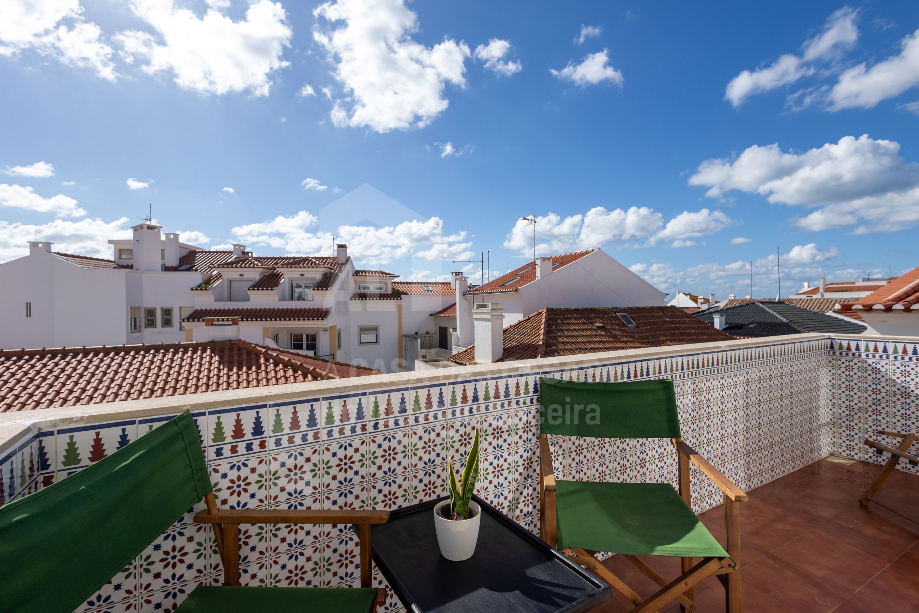 Terraço no telhado com planta em vaso, duas cadeiras verdes e parede de azulejos com padrão geométrico.