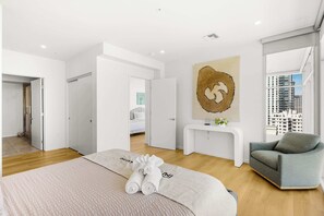 Quarto de hotel moderno com uma cama grande, uma poltrona cinza, um console branco e vista para a cidade.