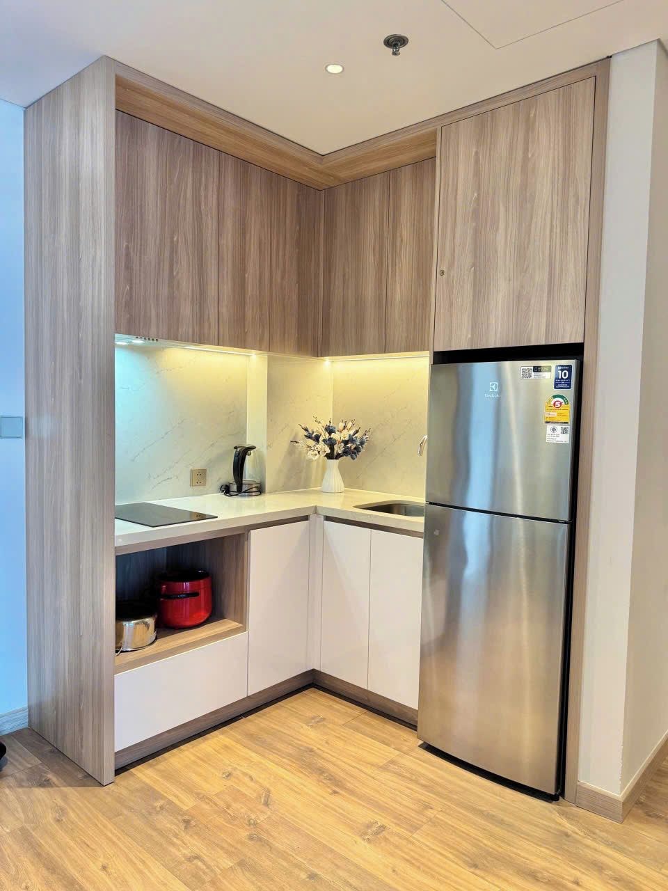 Cozinha compacta com geladeira de aço inoxidável, armários de madeira, bancada branca e forno embutido.