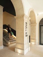 Um espaço arquitetônico imponente com altas colunas de pedra, uma escadaria e uma porta de vidro com a inscrição 