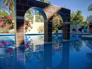 Piscina com água cristalina e piso ladrilhado, cercada por arcos de tijolo e plantas em vasos.
