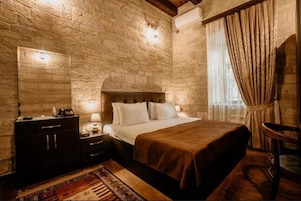 Quarto com parede de pedra, cabeceira de madeira, cama com roupa de cama branca, mesa de cabeceira com abajur e uma cadeira.