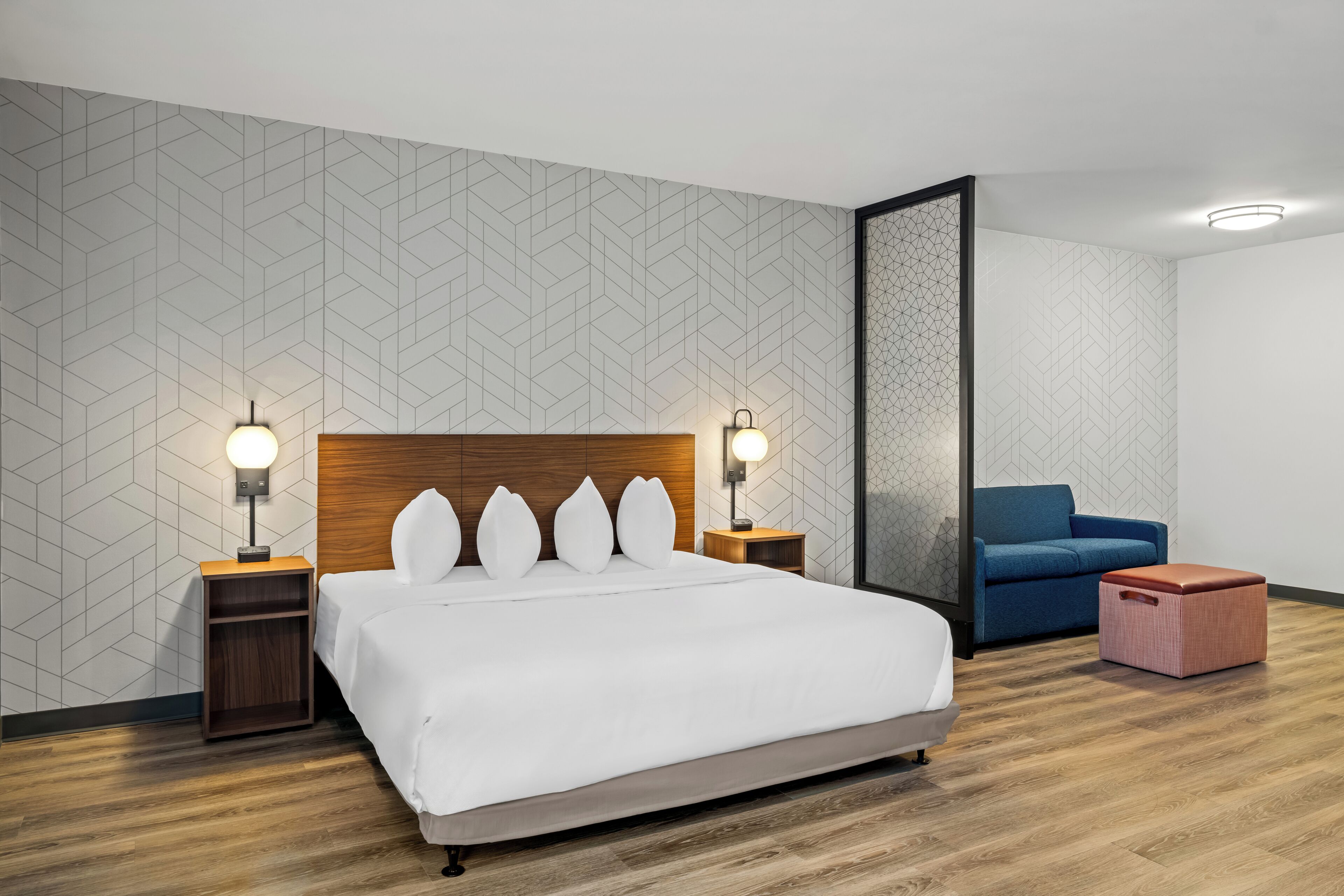 Quarto de hotel moderno com uma cama grande, duas mesas de cabeceira com abajures, um sofá azul e um banquinho de madeira.