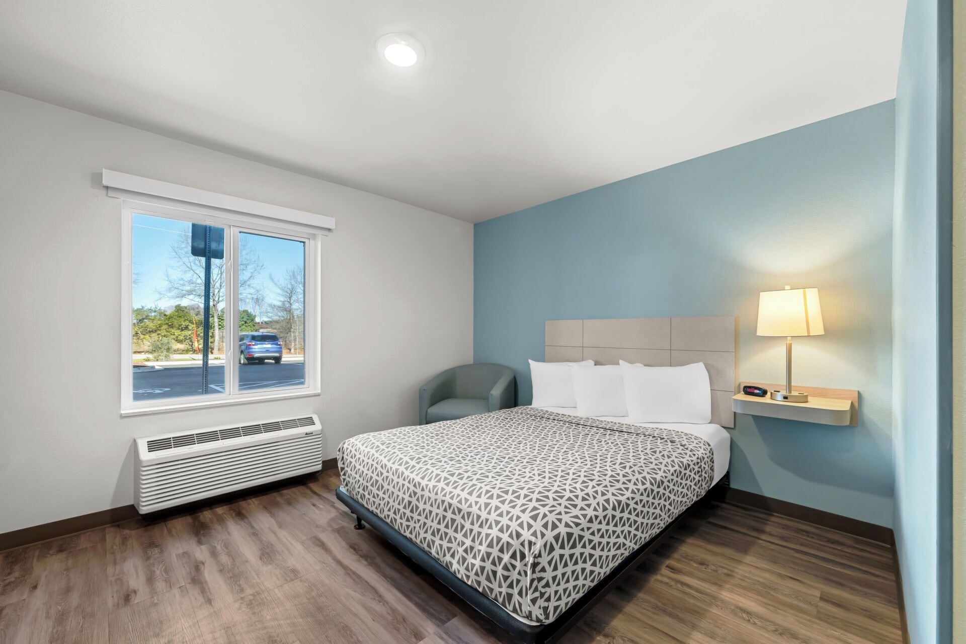Quarto de hotel moderno com uma cama grande, janela com vista para a rua, aquecedor portátil e mesa de cabeceira com abajur.