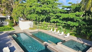 Uma piscina retangular com água cristalina, rodeada por um deck de madeira e espreguiçadeiras brancas.