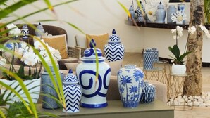 Diversos vasos de cerâmica azul e branca dispostos sobre uma mesa, com uma planta e um tronco de árvore de madeira ao fundo.