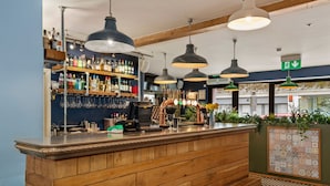 Uma área de bar com balcão de madeira, luminárias pendentes, prateleiras com garrafas e parede estampada.