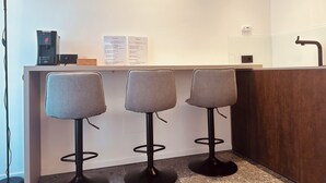 Uma área de bar moderna com três banquetas cinzentas, uma máquina de café e um quadro de menu.