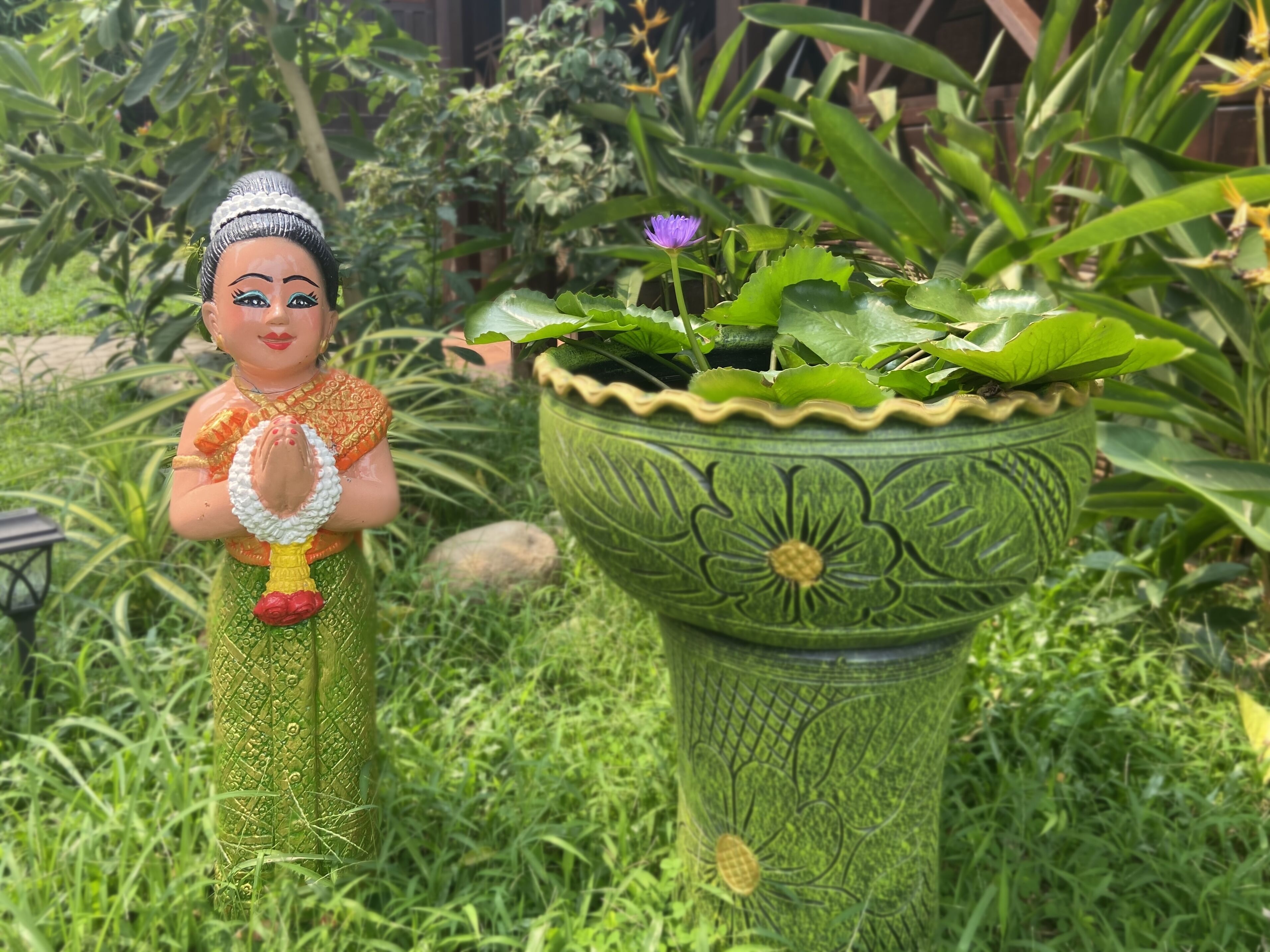 Estátua de uma figura vestida com traje tradicional segurando uma flor, ao lado de um vaso verde decorativo com uma flor em cima.