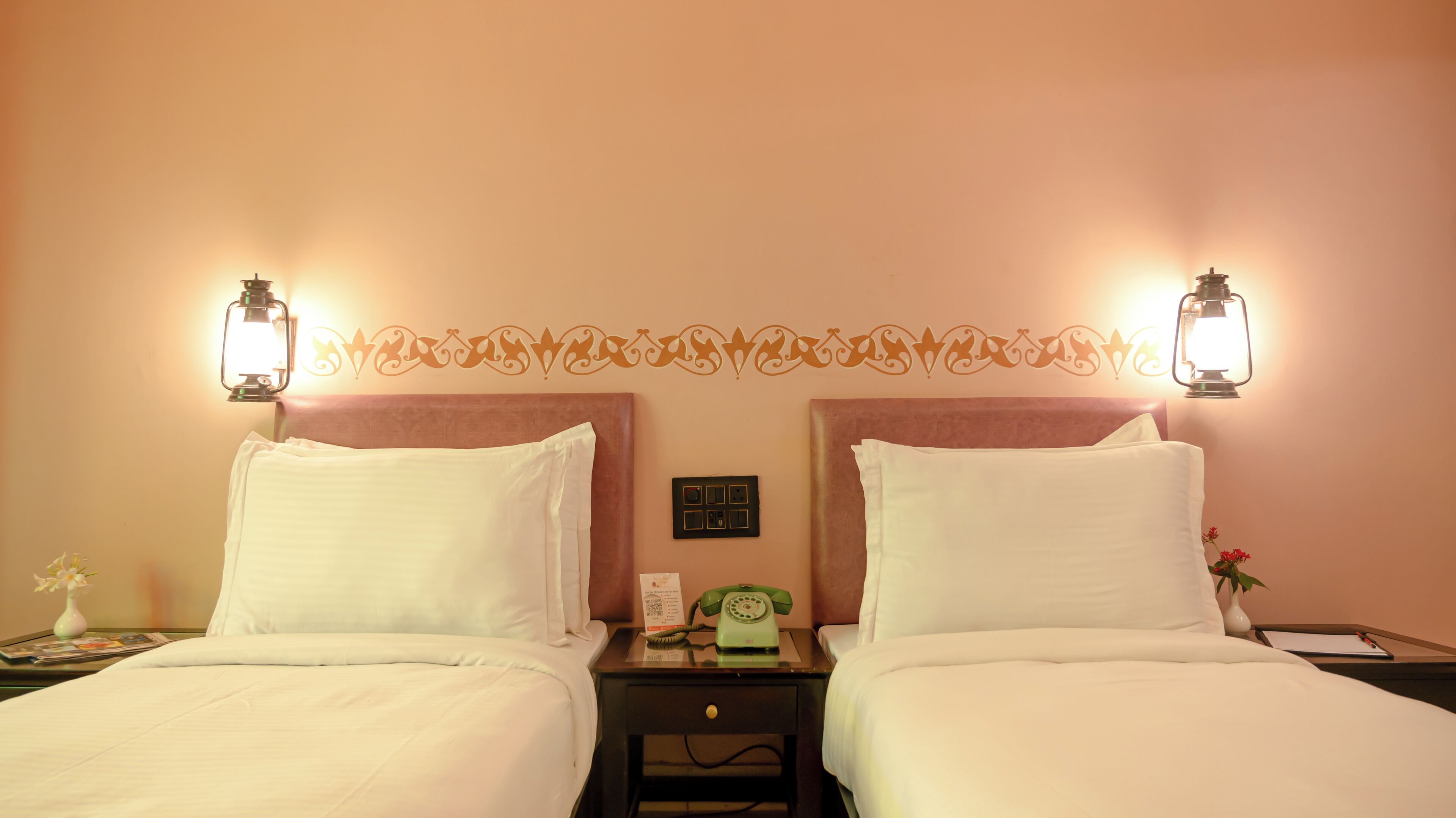 Quarto de hotel com duas camas, mesas de cabeceira, telefone e luminárias decorativas na parede.