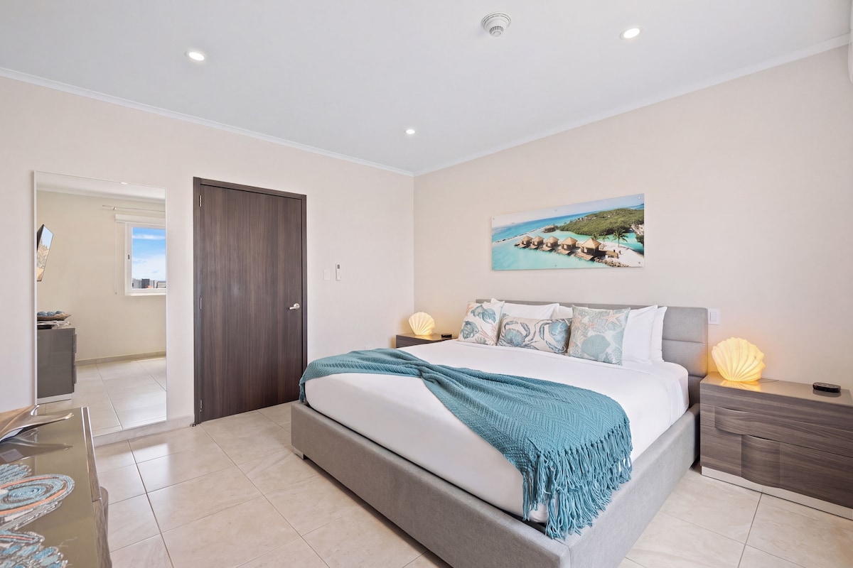 Quarto com cama, luminárias de cabeceira, um quadro e vista para o exterior.