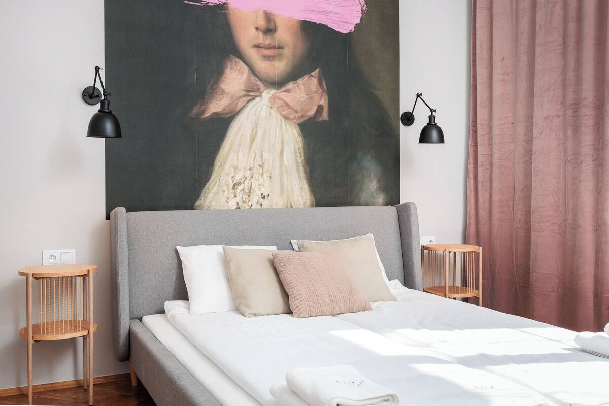 Cama bem arrumada com almofadas, um quadro grande na parede e duas mesas de cabeceira com luminárias.