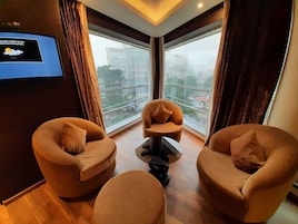 Quarto de hotel moderno com duas poltronas, uma mesinha de centro e uma televisão de tela plana na parede. Grandes janelas proporcionam vista para a paisagem urbana.