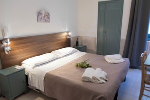 Cama bem arrumada com lençóis brancos e bege, mesa de cabeceira com garrafa e copos, criado-mudo pequeno e cabeceira de madeira.