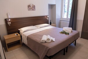 Uma cama bem arrumada com um espalha-cama marrom, travesseiros brancos e duas toalhas enroladas ao lado. Tem uma mesa de cabeceira com um telefone e uma garrafa de água. Um quadro está pendurado na parede acima da cama.