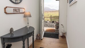 Quarto com uma mesa, uma lâmpada, um letreiro com a palavra 'FAILTE' e vista para uma paisagem rural através de uma porta aberta.