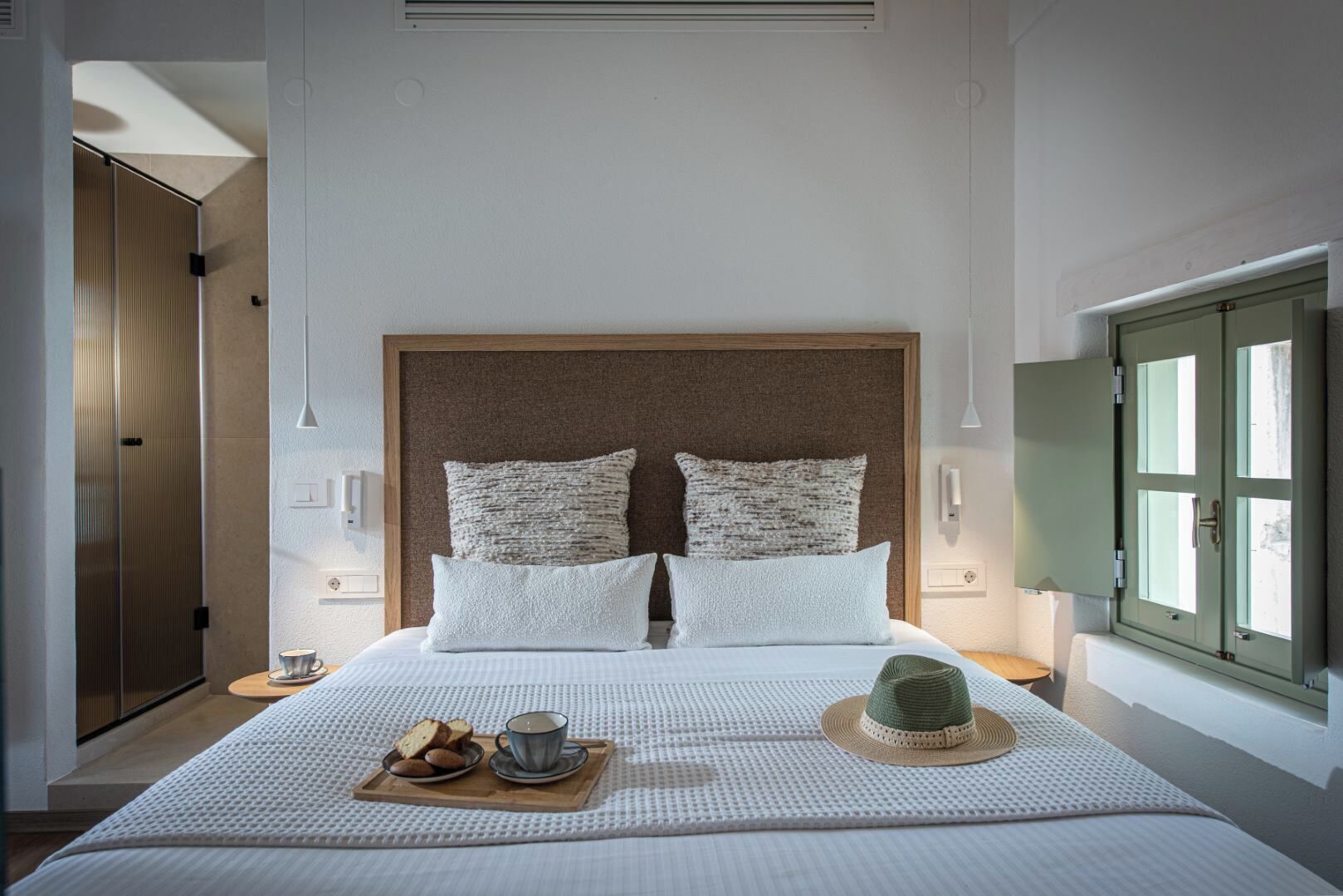 Cama bem arrumada com almofadas, bandeja com pão e uma chávena, e um chapéu sobre a cama.
