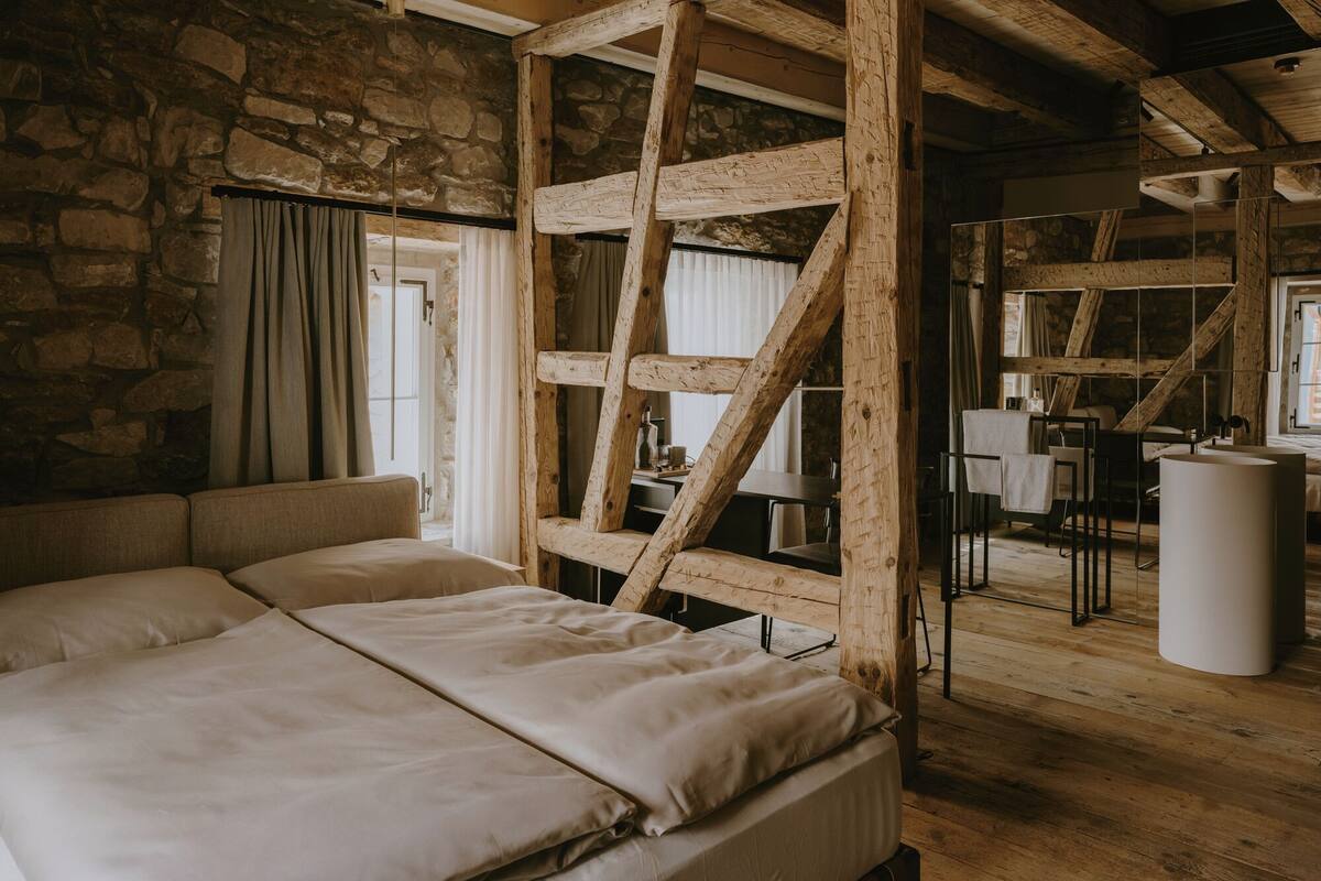 Quarto rústico com vigas de madeira aparentes, paredes de pedra, uma cama, uma mesa e uma cadeira.
